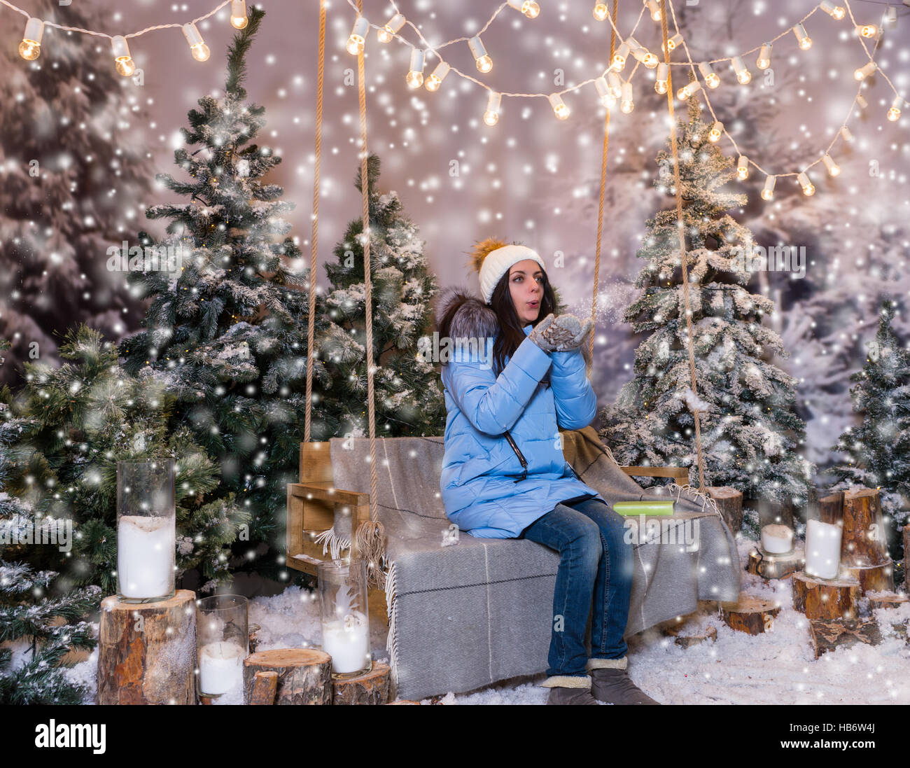 Woman in blue down jacket blows snowflakes while sitting on a swing ...
