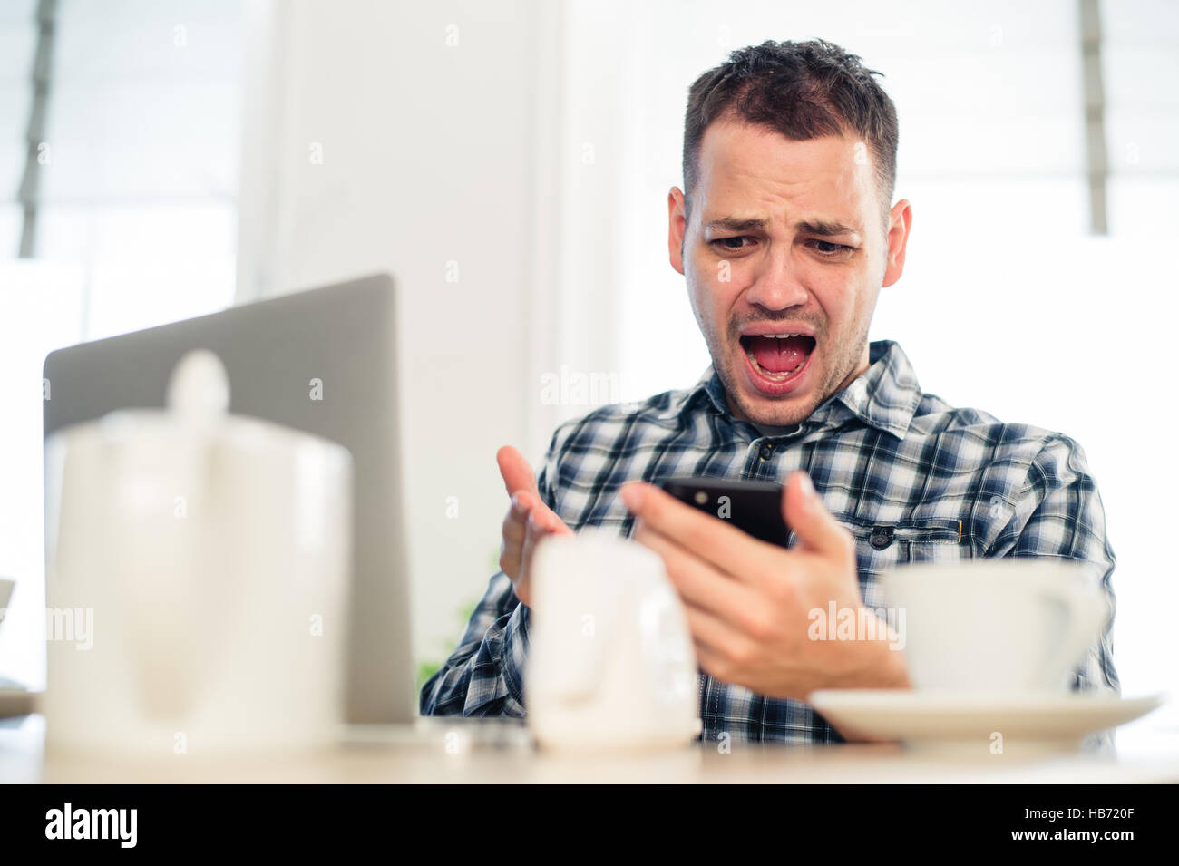 Bad news. Angry young man holding a mobile phone and grimacing at cafe ...