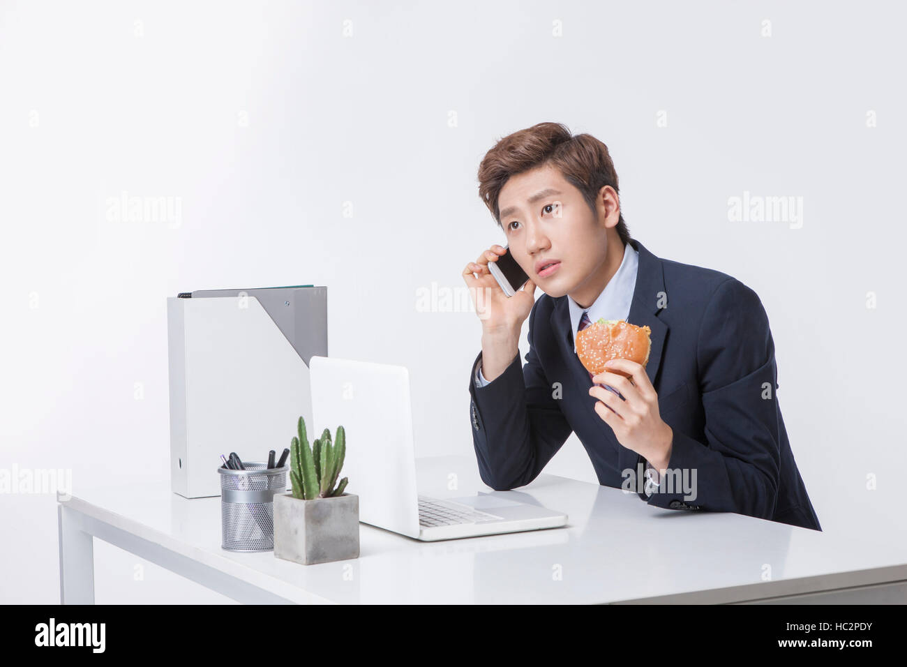 Young Korean businessman talking on cellphone eating hamburger at work ...