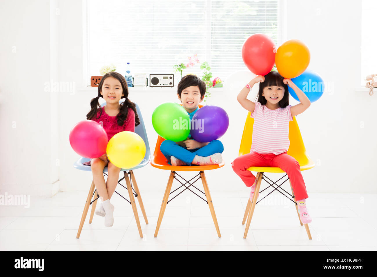 Three smiling children with balloons sitting on chairs Stock Photo - Alamy