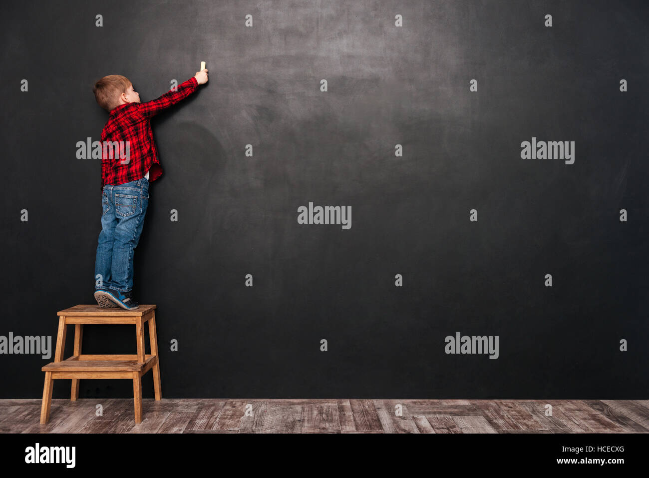 Photo of a little cute child standing on stool near blackboard and ...
