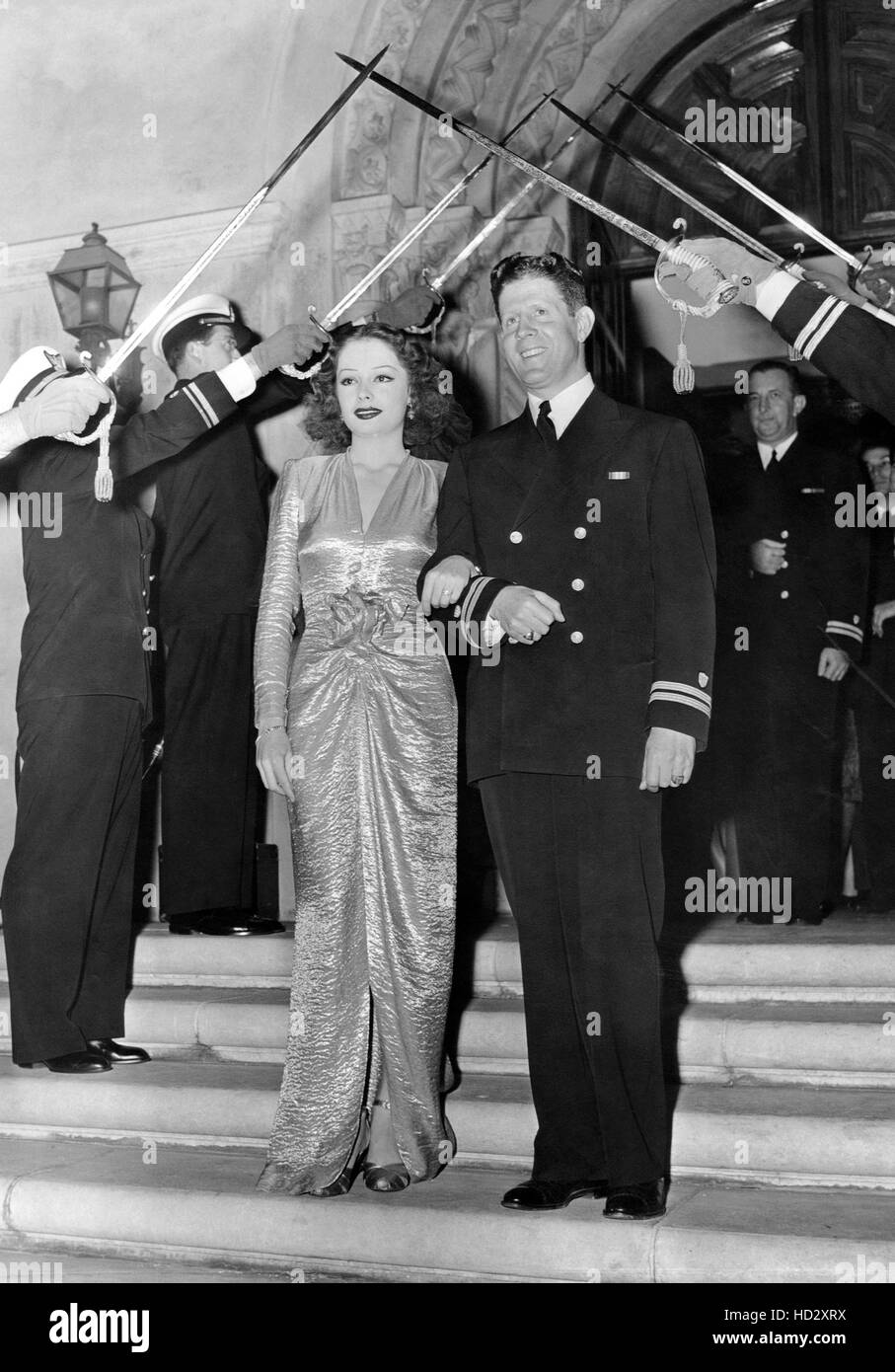 From left: Newlyweds Jane Greer, Rudy Vallee, walking under arc of ...