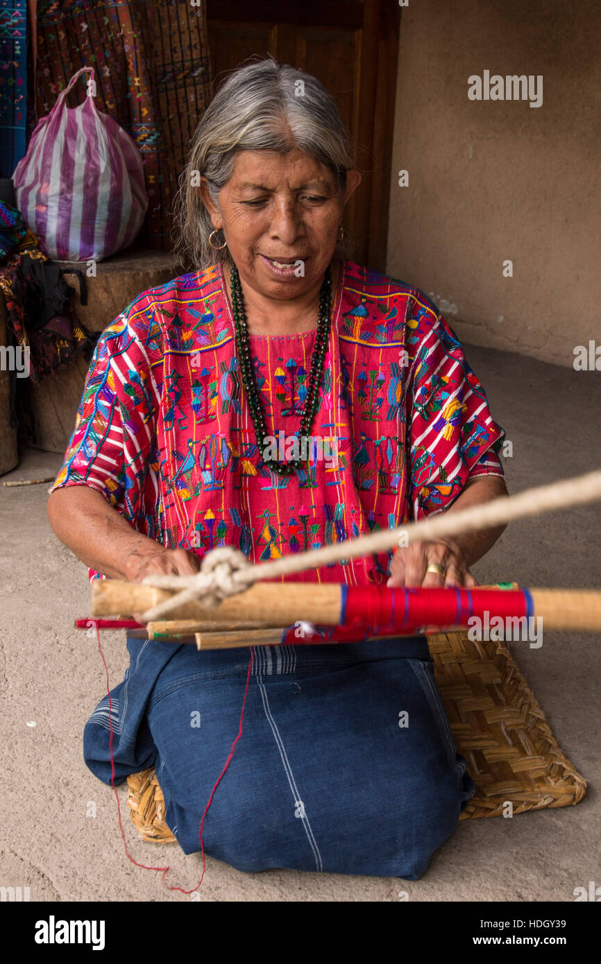 An older, grey-haired Mayan woman weaves fabric on a backstrap loom ...