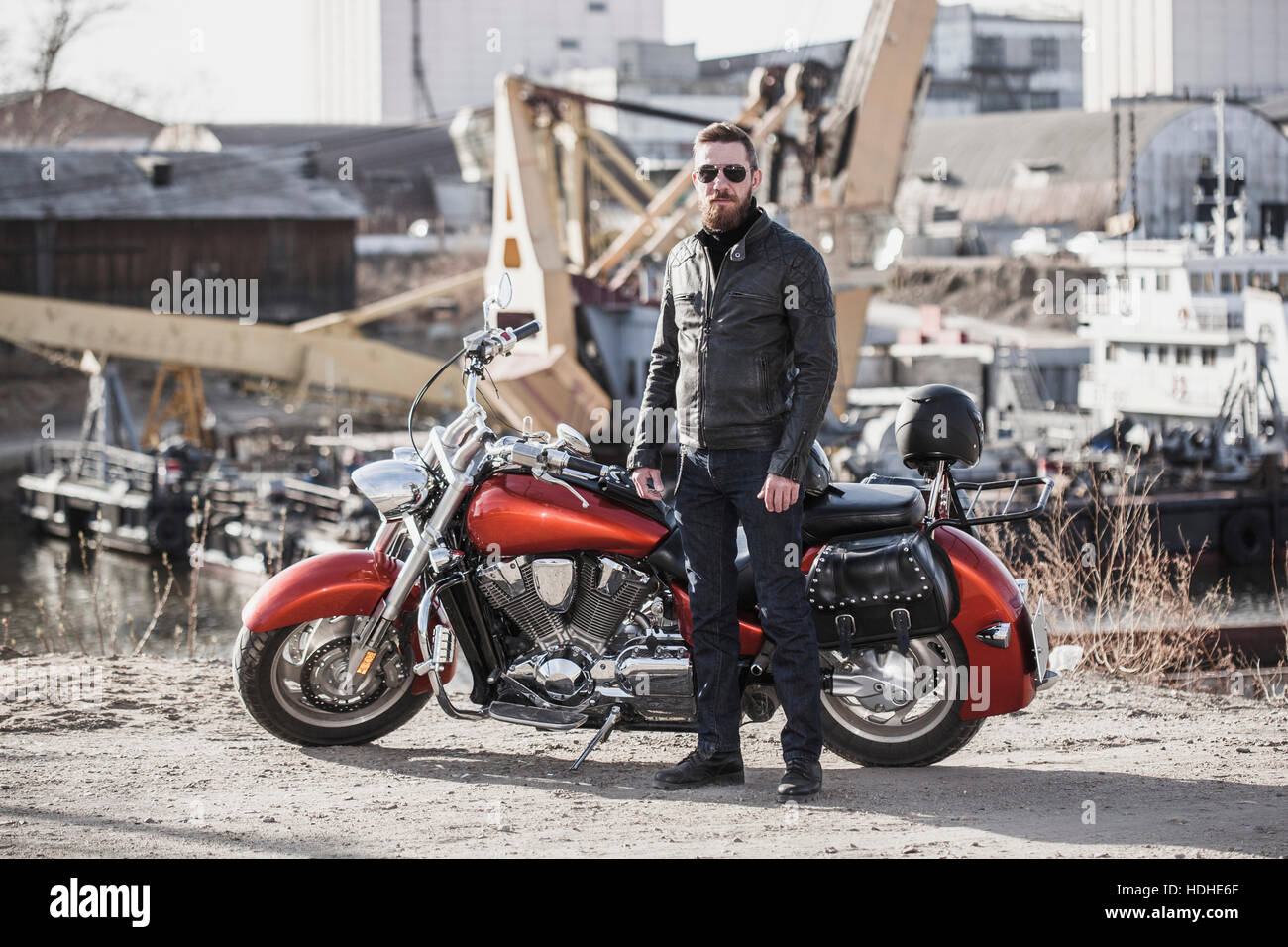 Full length portrait of biker standing by motorcycle against industrial ...