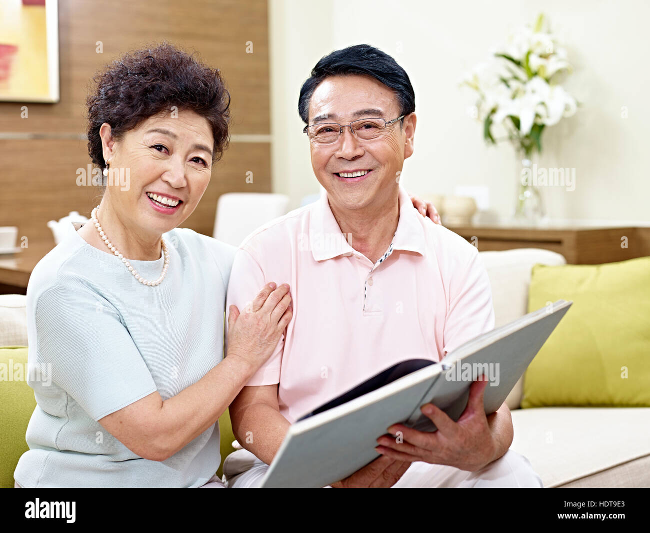 senior asian couple sitting on couch looking at camera with book in hands, happy and smiling ...