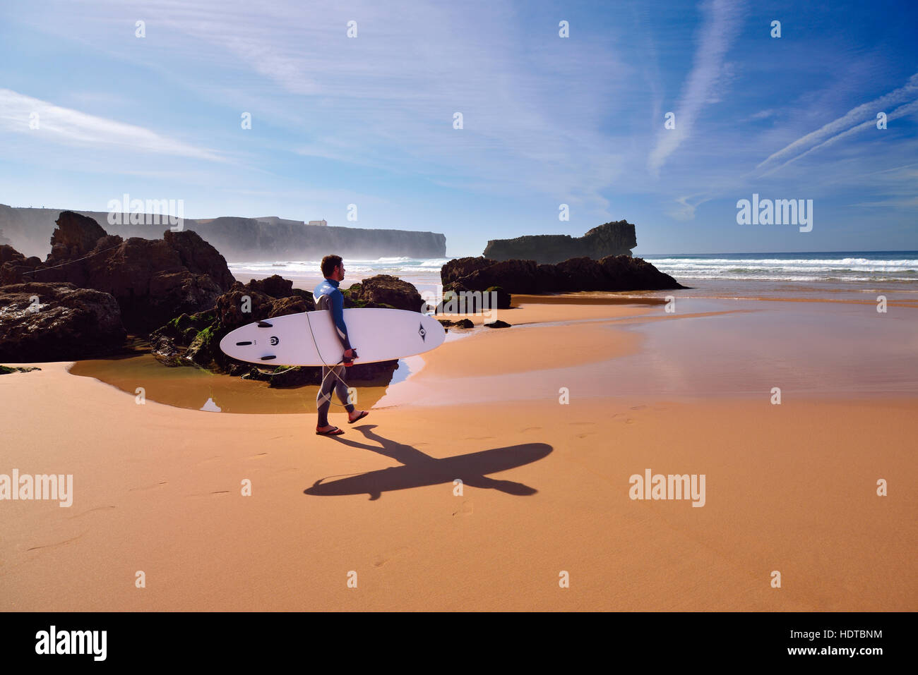 Surfer walking with his board towards the ocean at rocky beach with ebb ...