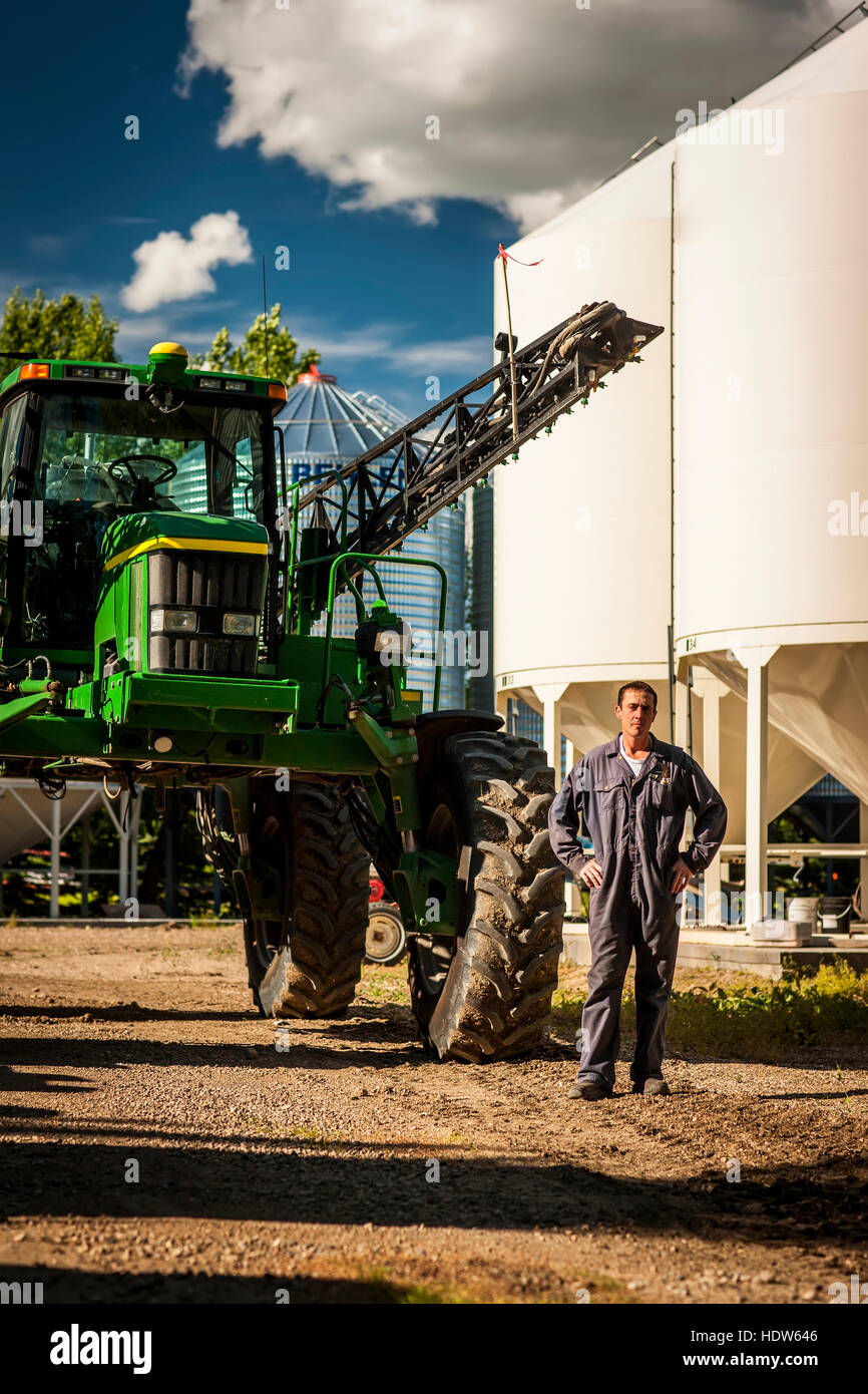 A young farmer stands posing beside a large tractor and grain storage ...