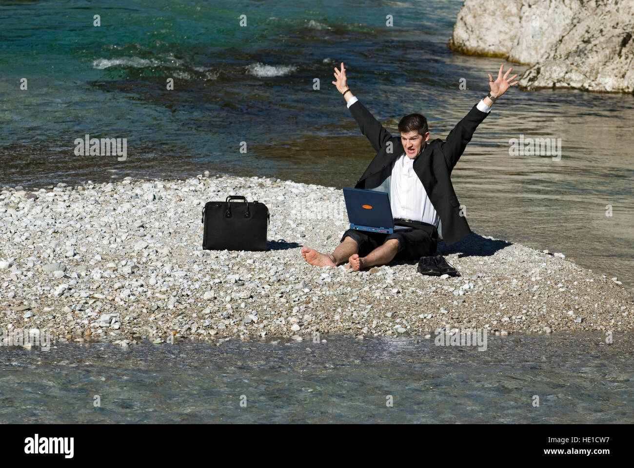 Businessman sitting at a stream's edge, working on his laptop outdoors ...