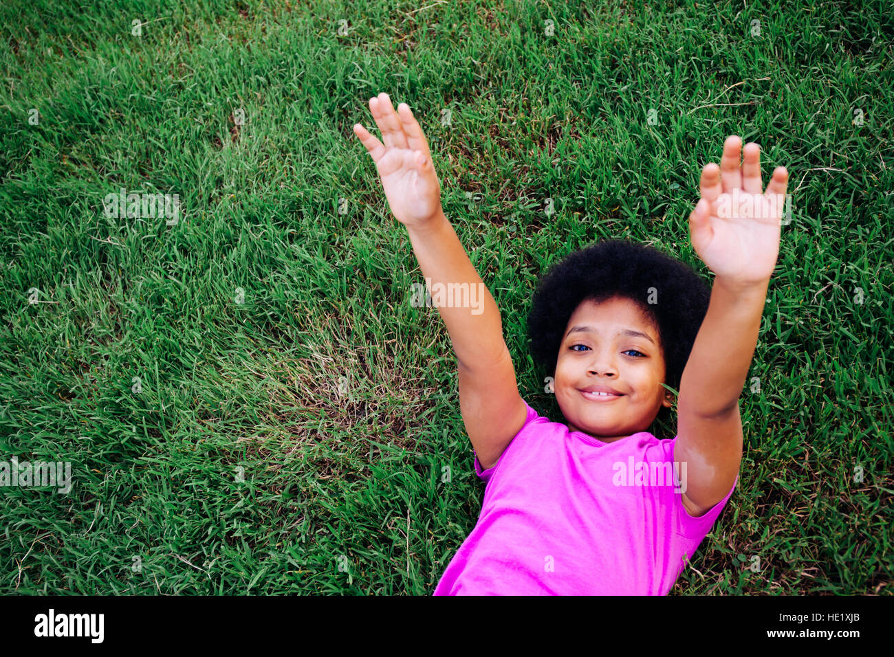 Little kid raising hands up and laying down on green grass in the park ...