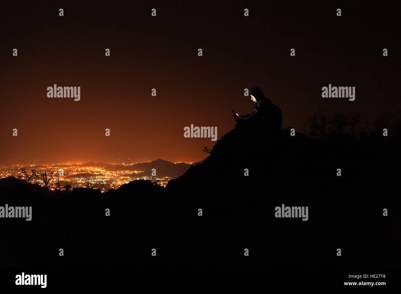 Young man with phone on top of the hill observing the night city view ...