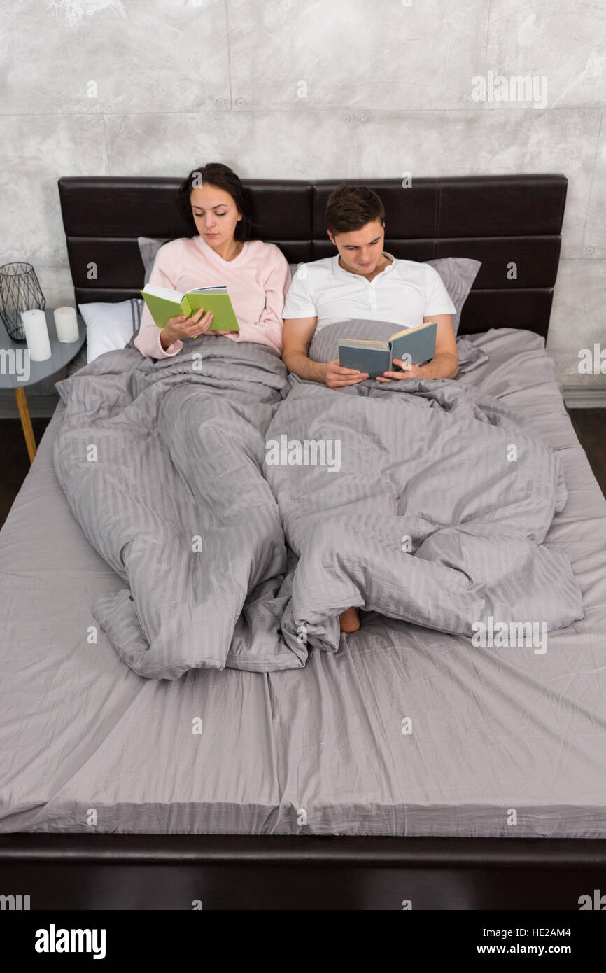Young attractive pair reading books while lying in the bed and wearing ...