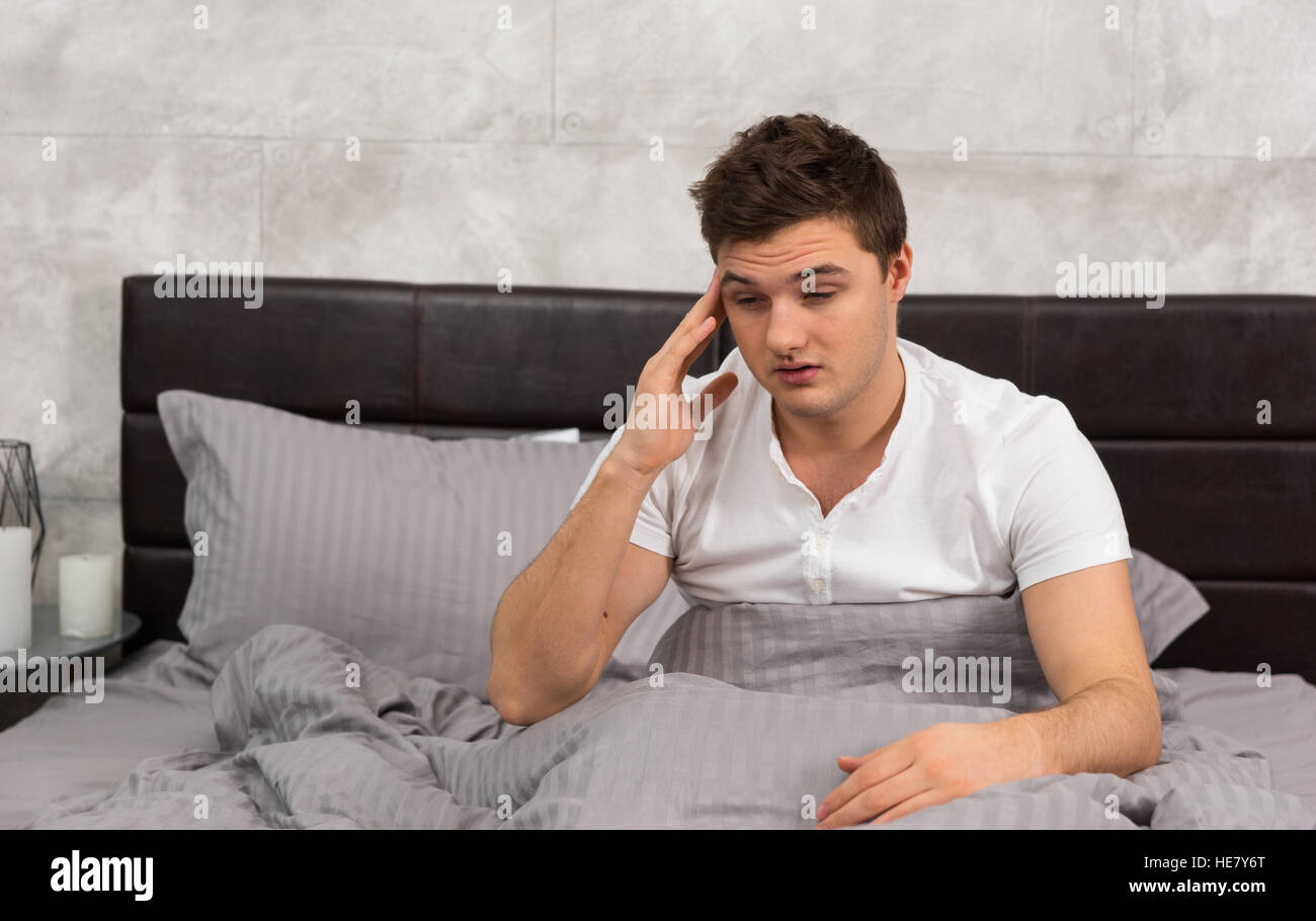 Young man woke up with hangover sitting in stylish bed with grey colors ...