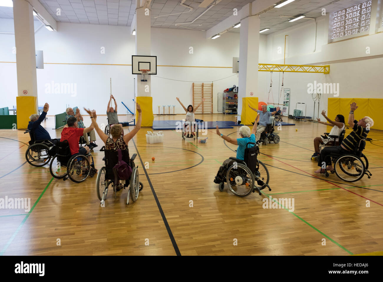 An exercise class in a sports hall for wheelchair users lead by a woman ...
