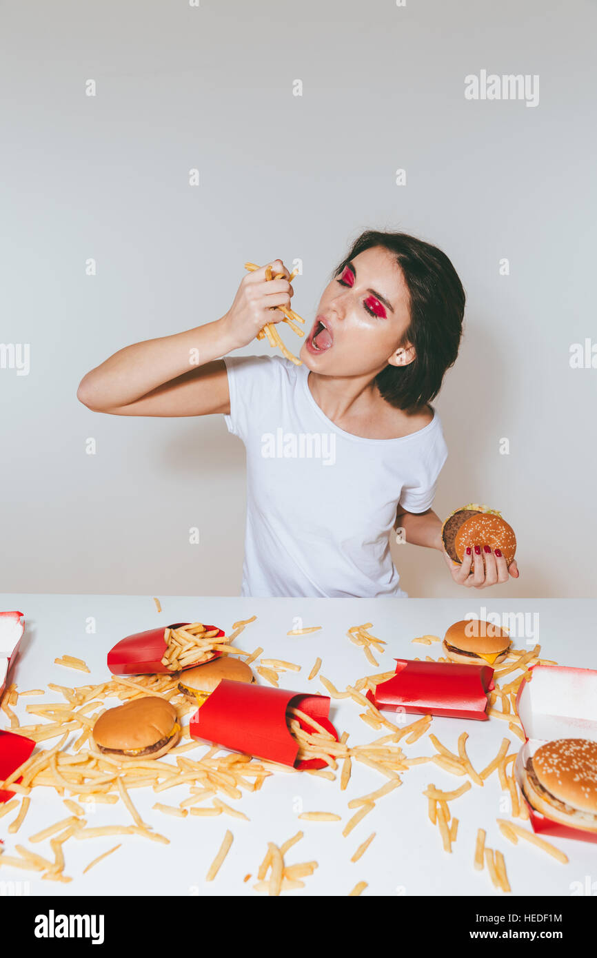 Pretty young woman sitting and eating french fries at the table with ...