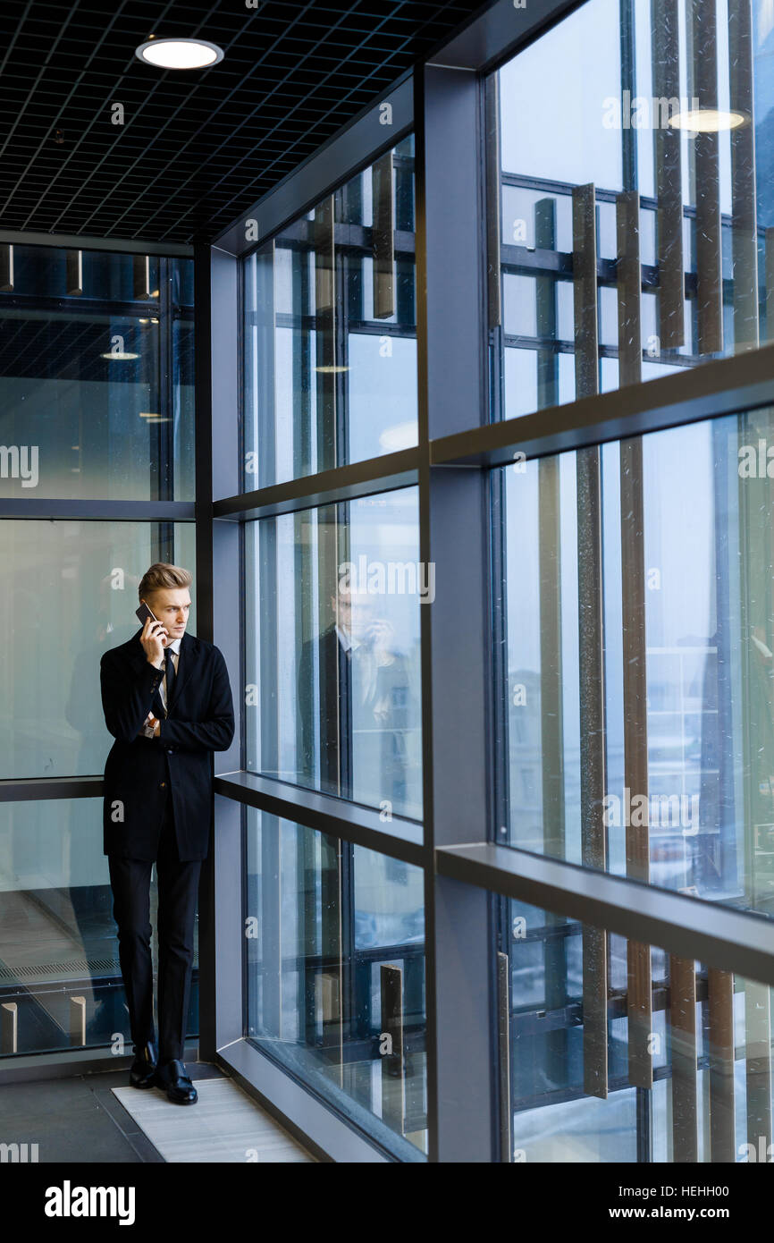 Young man standing by window of office-center and talking by cellphone ...