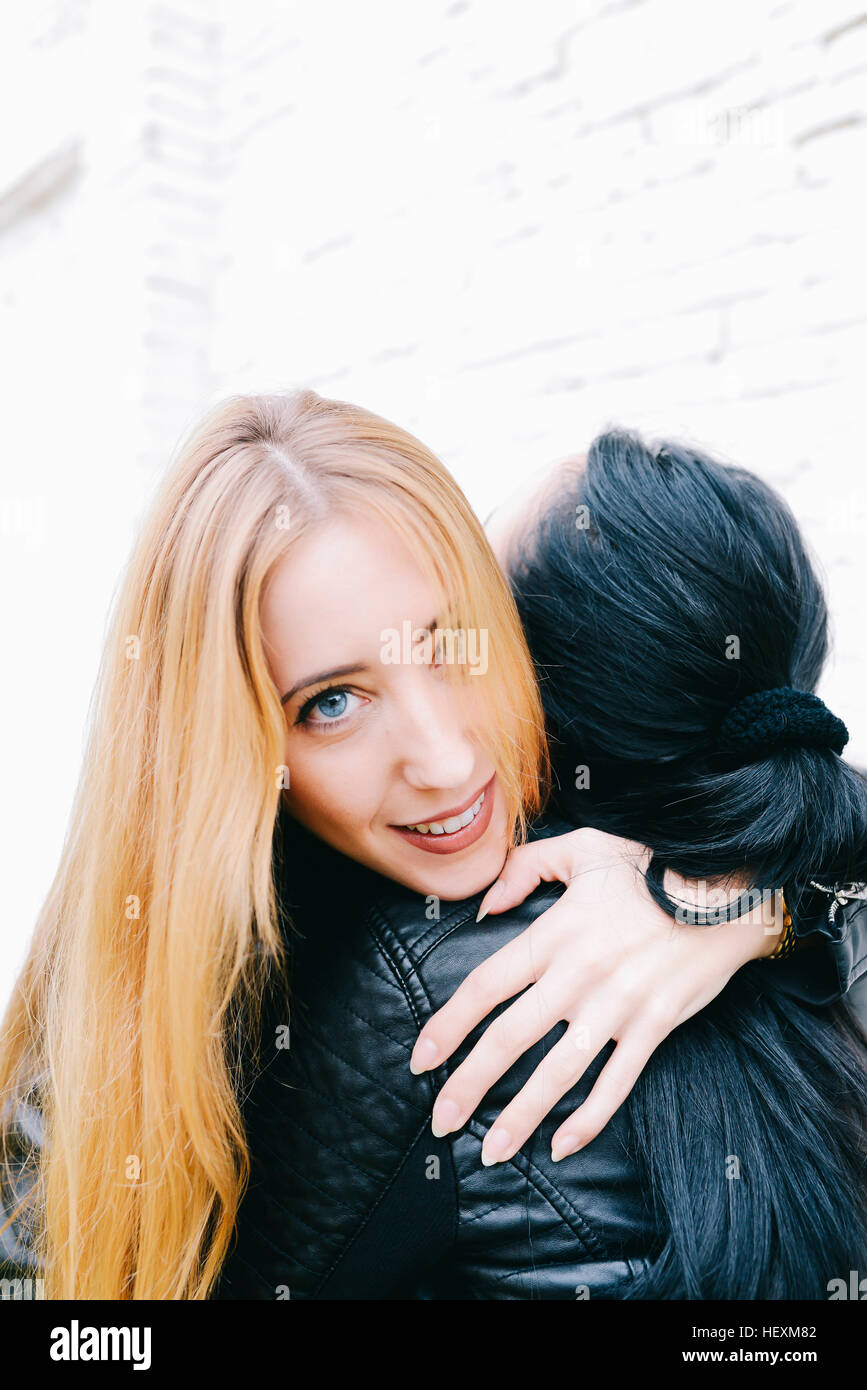 Portrait of young woman hugging her friend in front of white brick wall ...