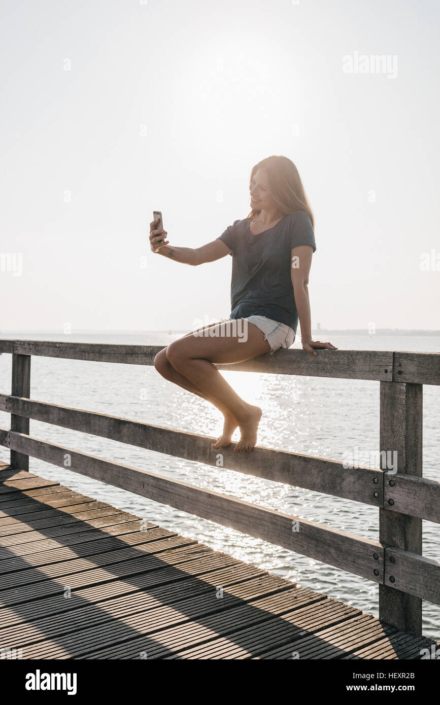 Young woman sitting on jetty at backlight taking selfie with cell phone ...