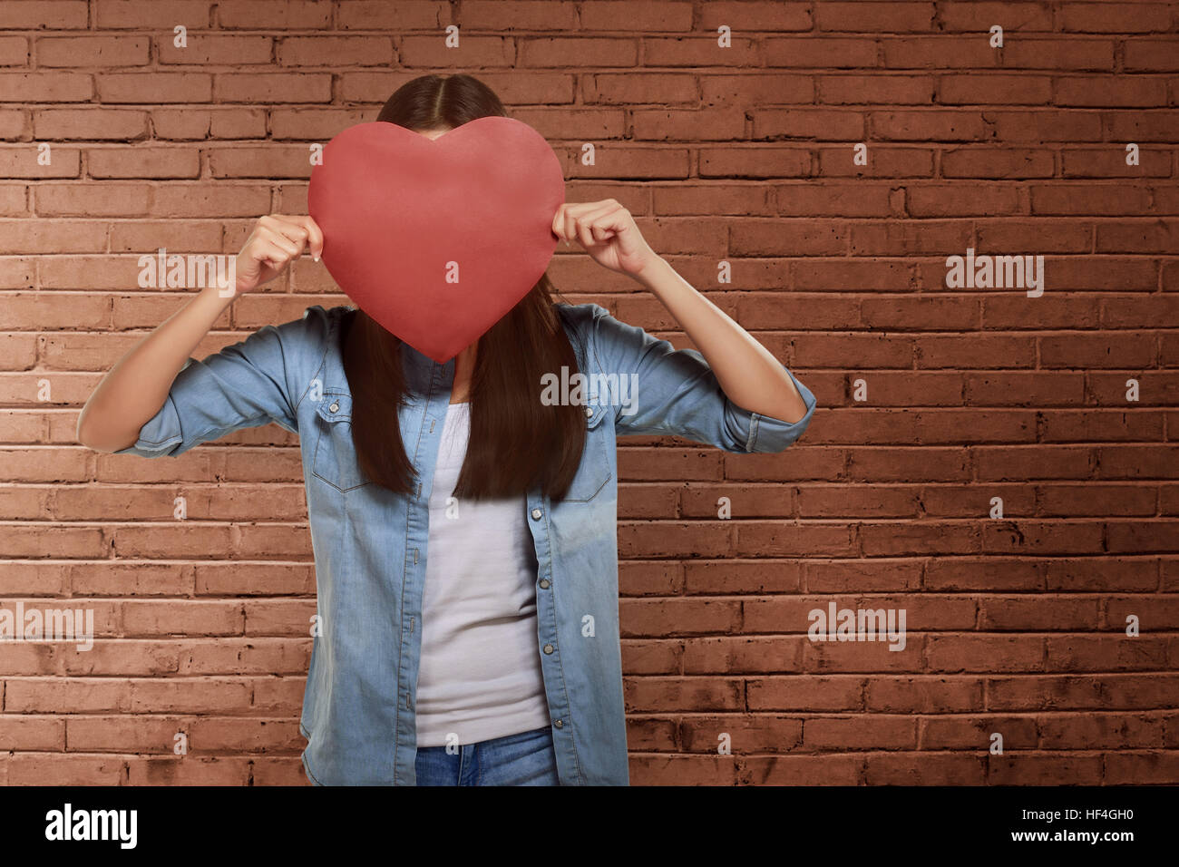 Asian girl holding heart shape over her face on the brick wall ...