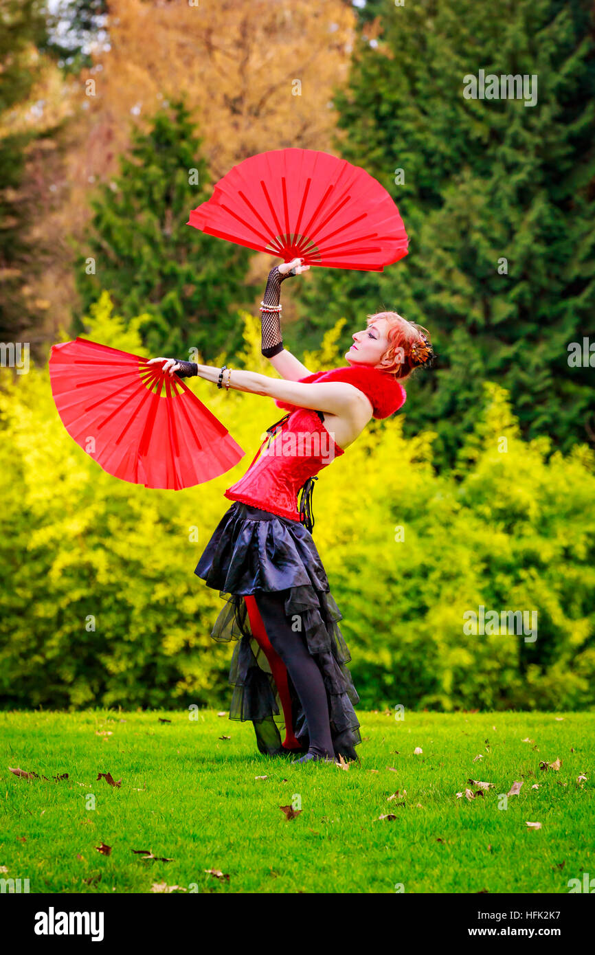 Young beautiful woman in circus costume play with red fans in the park ...