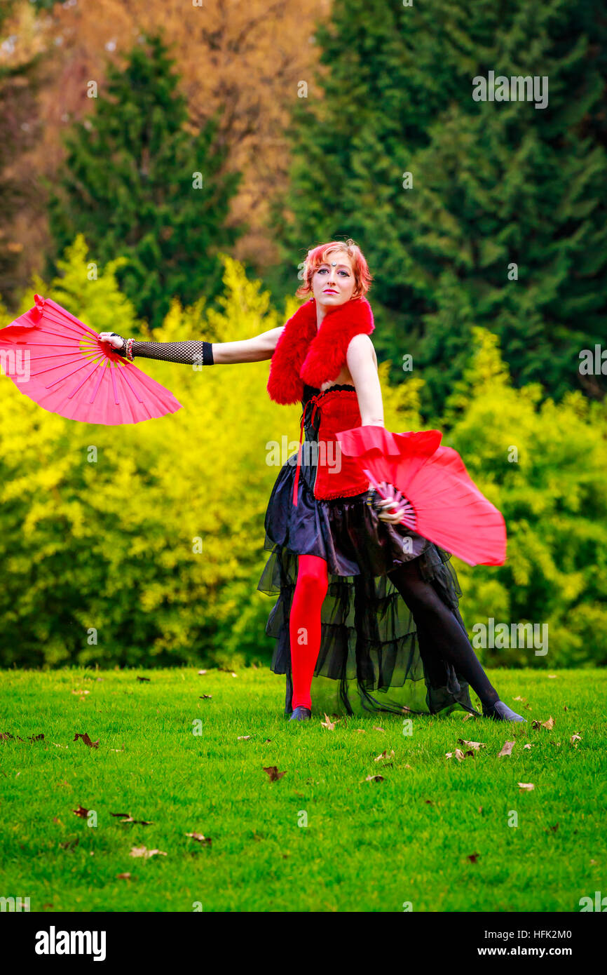 Young beautiful woman in circus costume play with red fans in the park ...