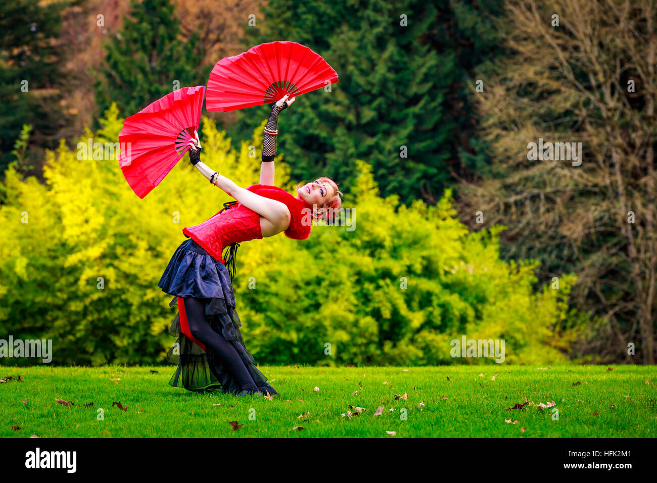 Young beautiful woman in circus costume play with red fans in the park ...