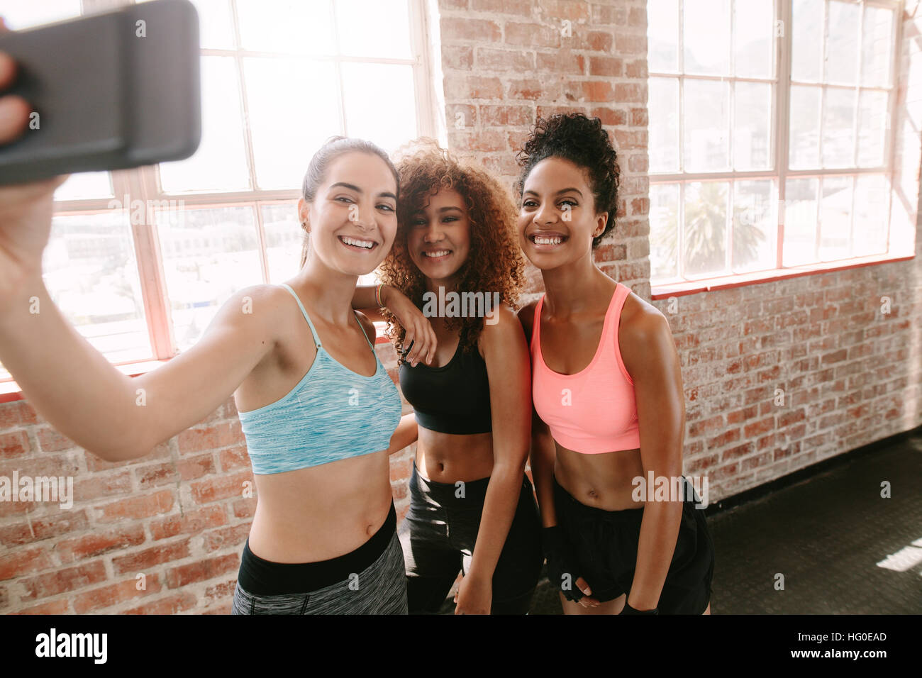 Group of happy young women taking selfie by smartphone in gym. Multiracial group of girls in ...