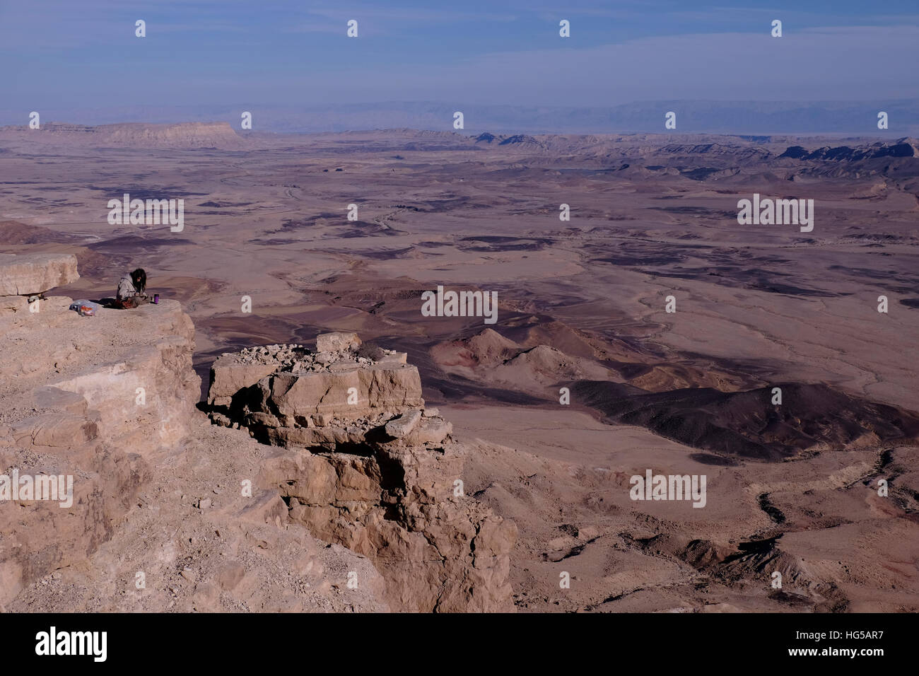 A lone female traveler sits on a cliff rim at the edge of Mount Ramon ...
