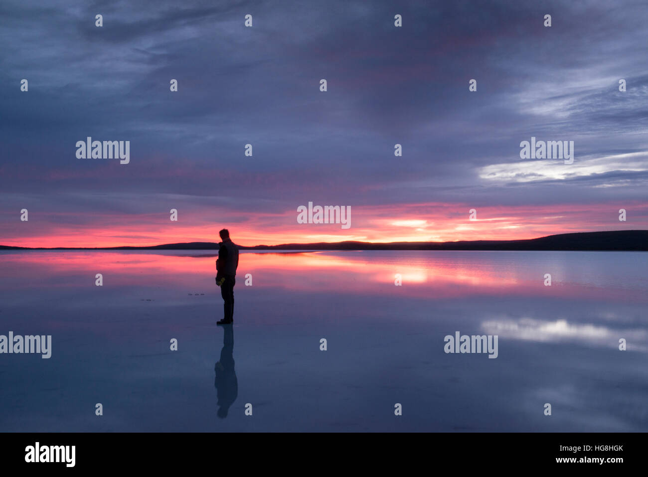 A silhouette of a man stands on a reflective lake in sunset or sunrise ...