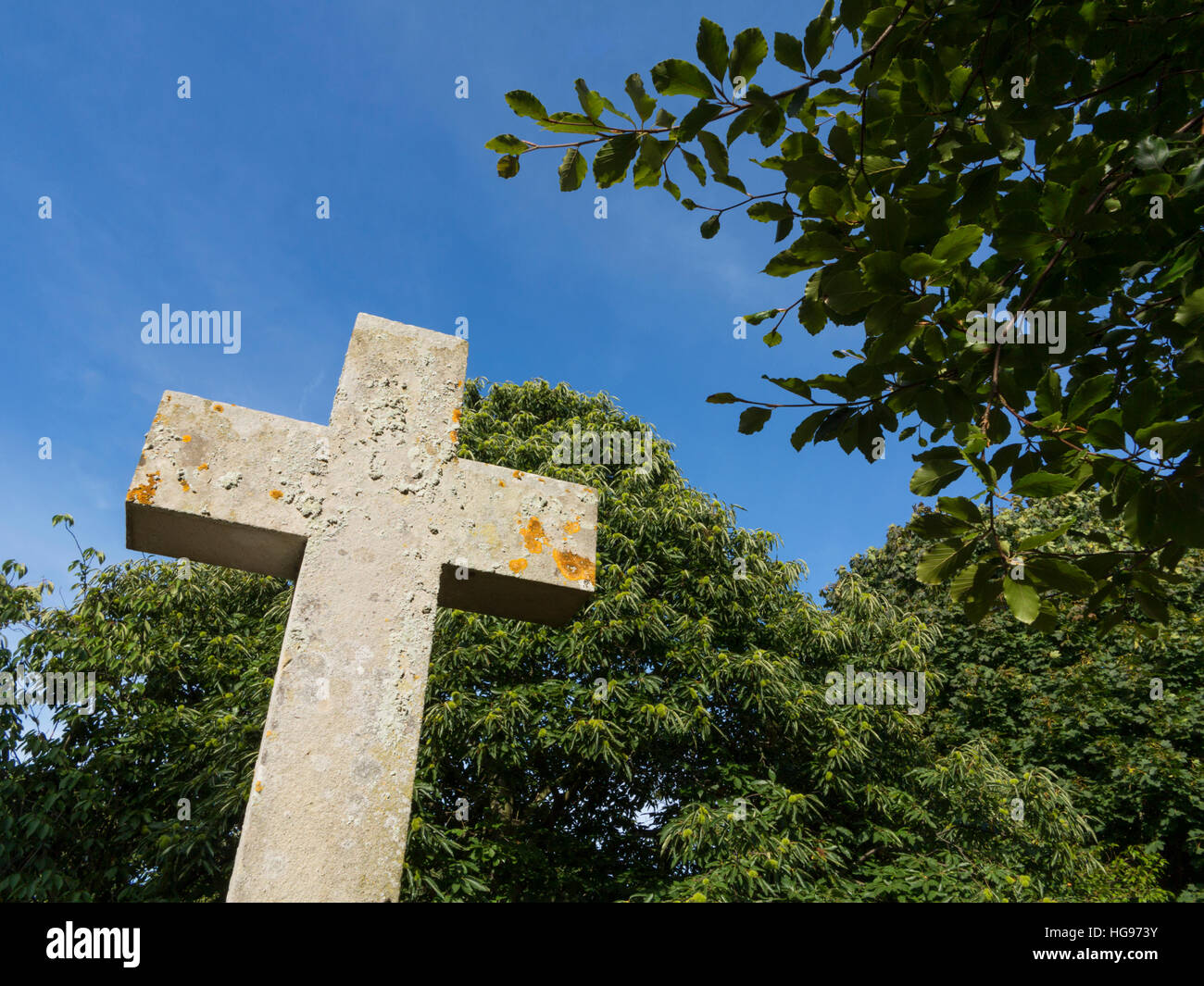 A single plain limestone cross headstone against a blue sky and trees ...