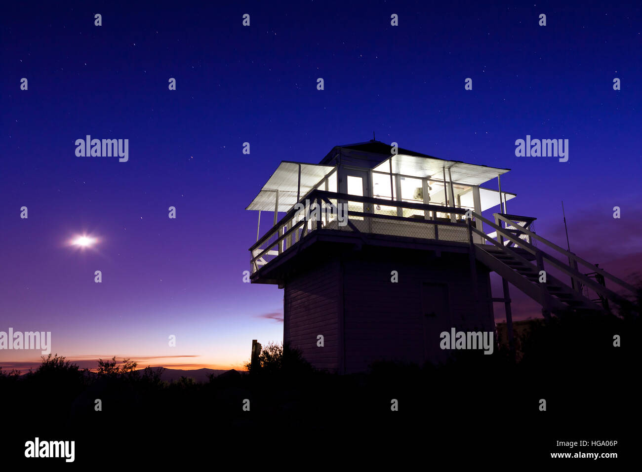 Lit Fire lookout at night with sunset and moon in the sky Stock Photo ...