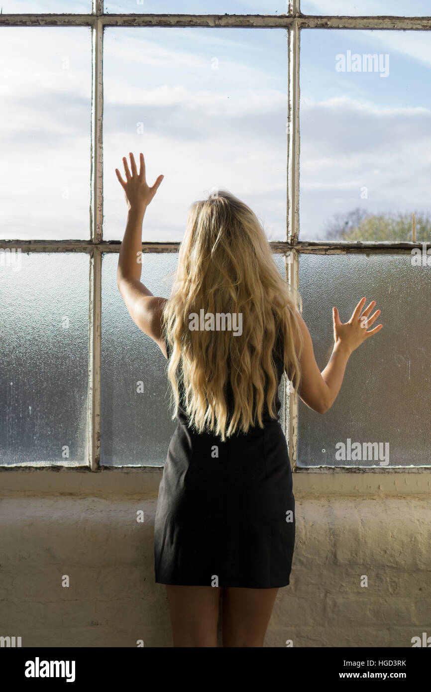 Rear view of a blond woman standing by the window hands touching the ...