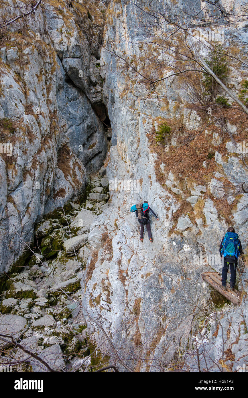 Two people climbing a difficult trail along a cliff face towards a cave ...