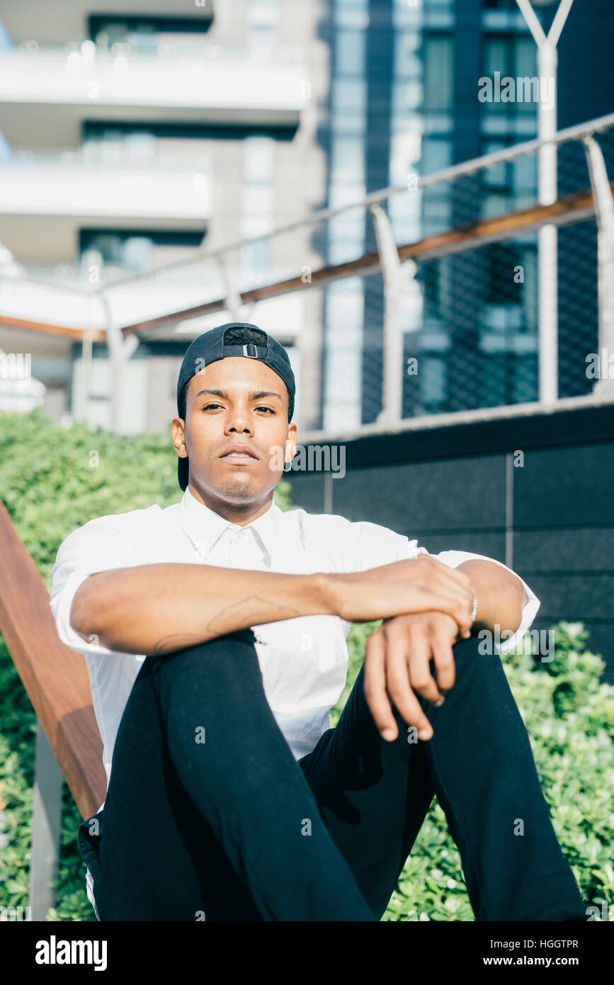 Young handsome american african man sitting outdoor in city back light, looking at camera ...