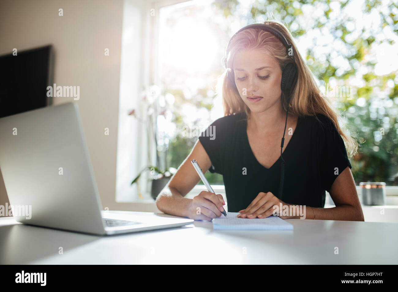Indoor shot of woman taking notes in kitchen. Female wearing headphones ...