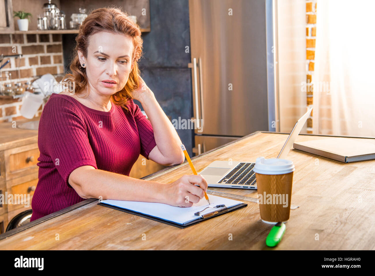 Woman making notes sitting in kitchen with laptop Stock Photo - Alamy