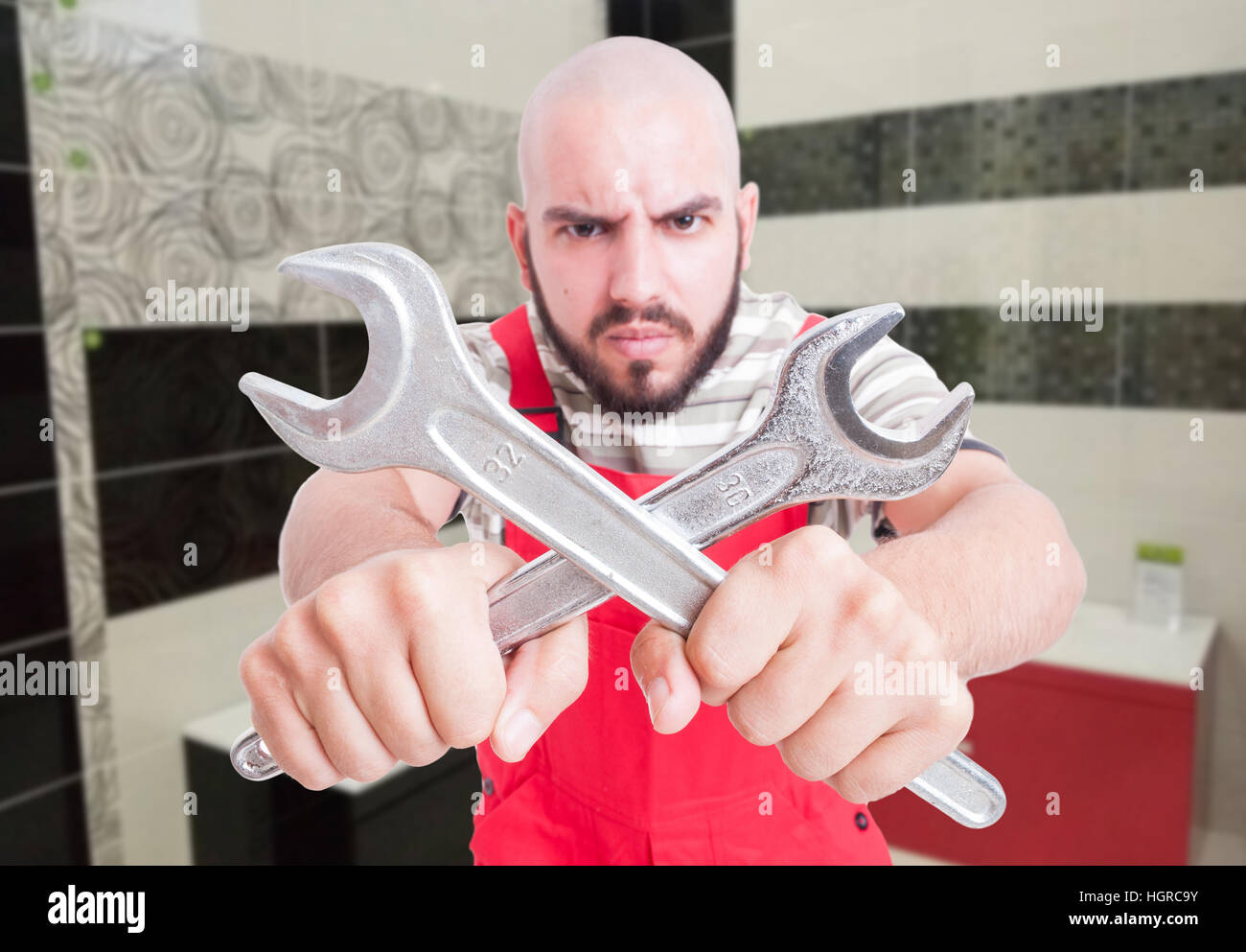 Angry plumber in red overalls holding crossed wrenches in the batchroom ...