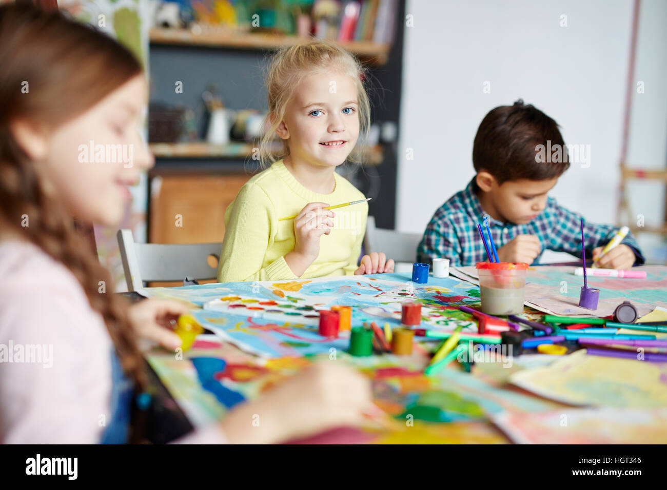 Little girl with paintbrush looking at camera among her classmates ...