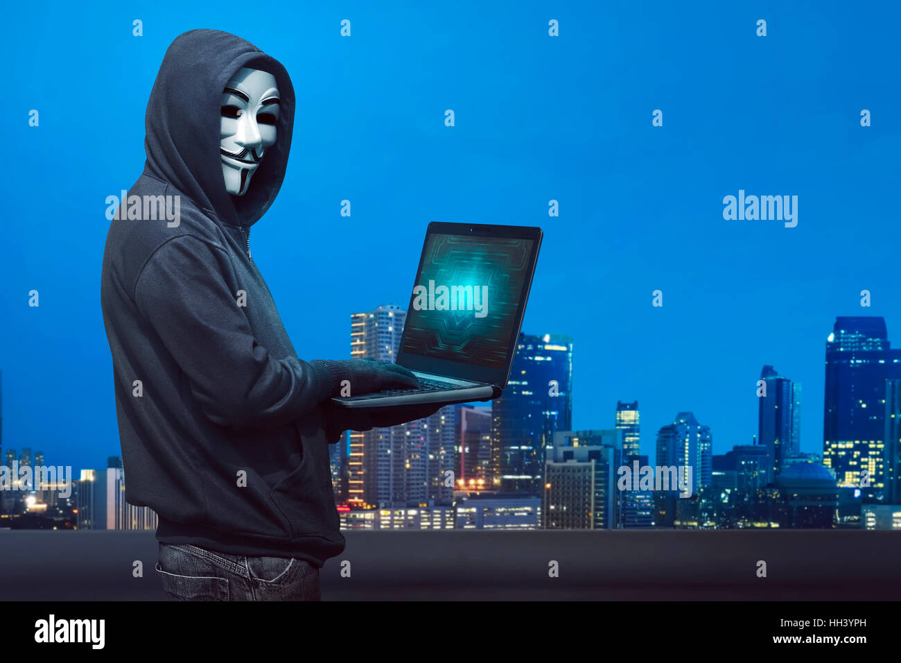 Hooded hacker with anonymous mask typing on laptop against modern city ...
