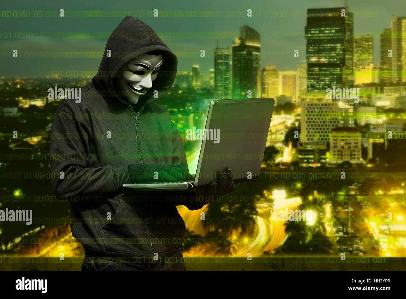 Hacker man with mask typing on laptop against modern city background ...