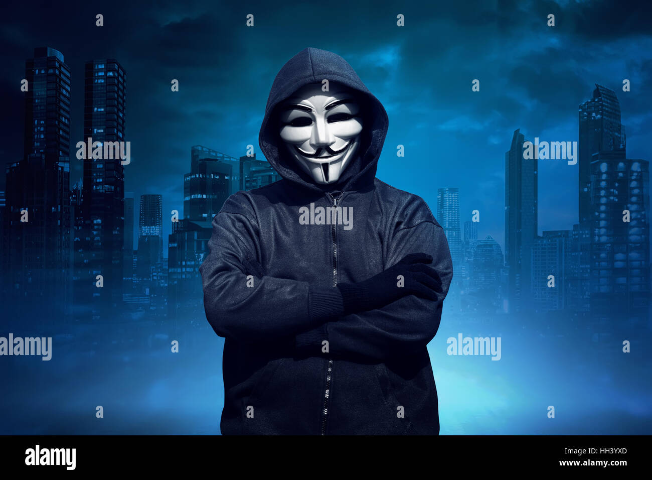 Hooded man with anonymous mask standing against cityscape background ...