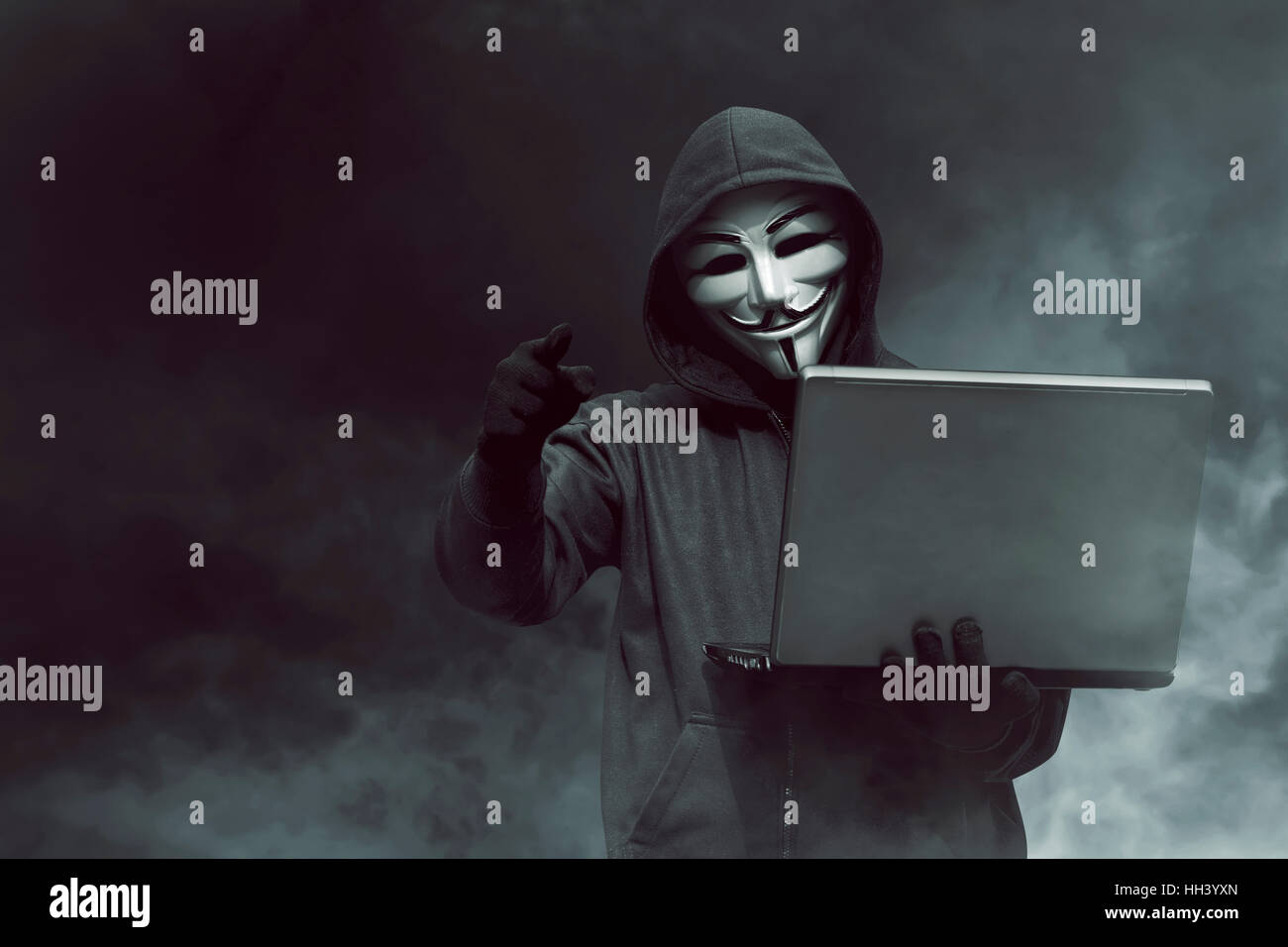 Portrait of hooded hacker with mask holding laptop while standing ...