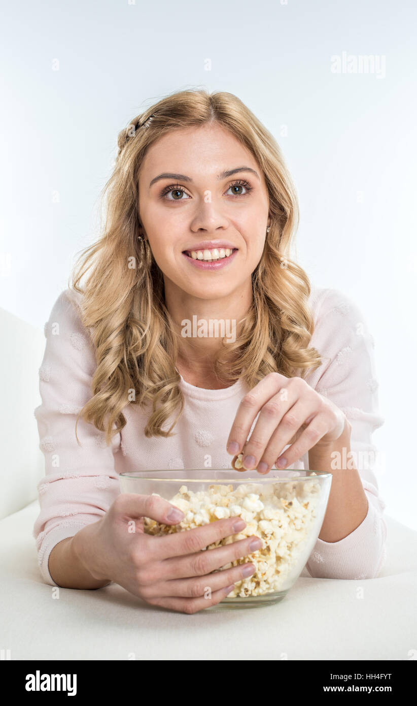 Smiling blonde woman eating popcorn from glass bowl and looking away ...