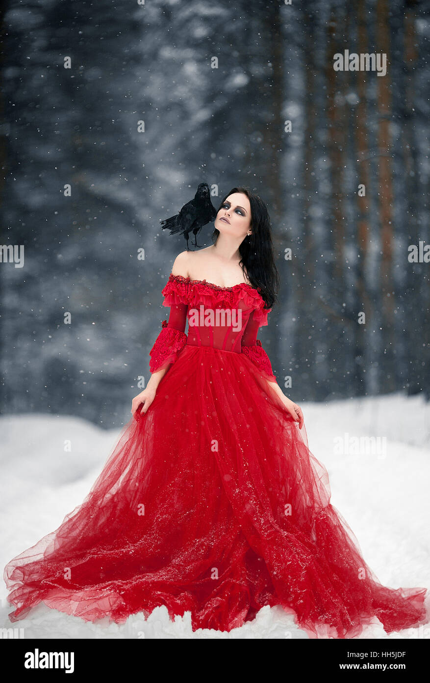 Woman witch in red dress and with raven on her shoulder in snowy forest ...
