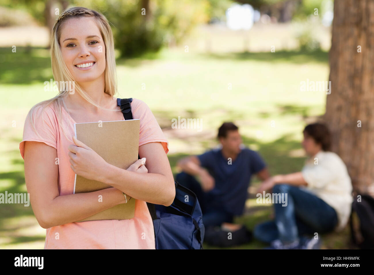 Portrait of a pretty girl smiling while holding a textbook in a park ...