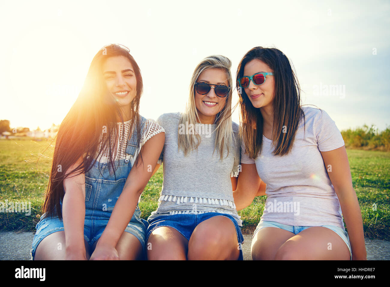 Portrait of three young female friends in summer wears sitting together against sunny landscape ...
