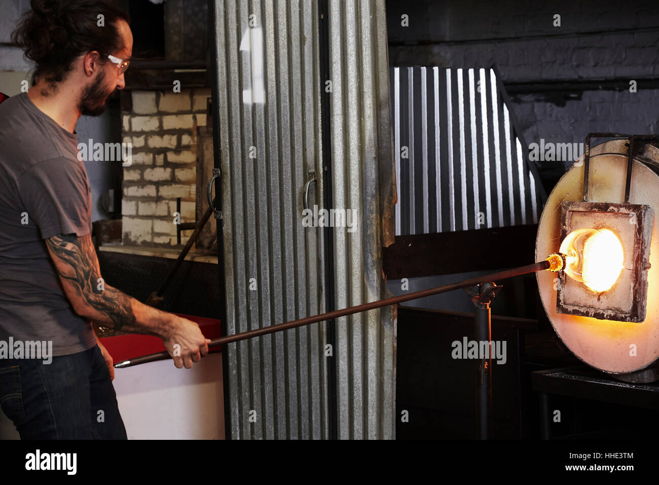 A glassblower holding a piece of molten glass to the furnace to heat it ...