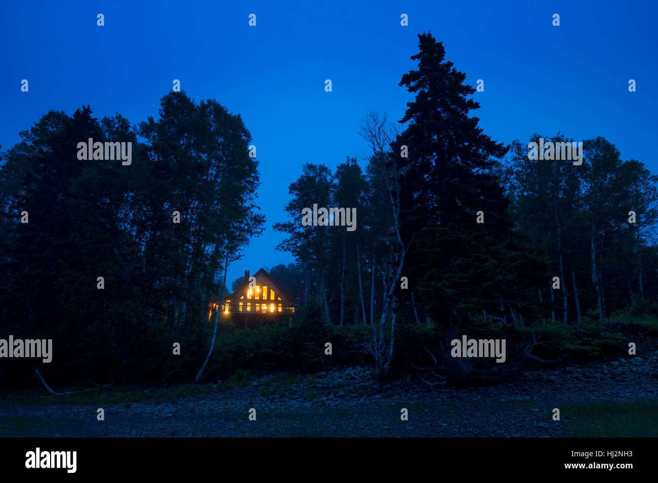 A log cabin is lit up at dusk along a tree covered shore Stock Photo ...