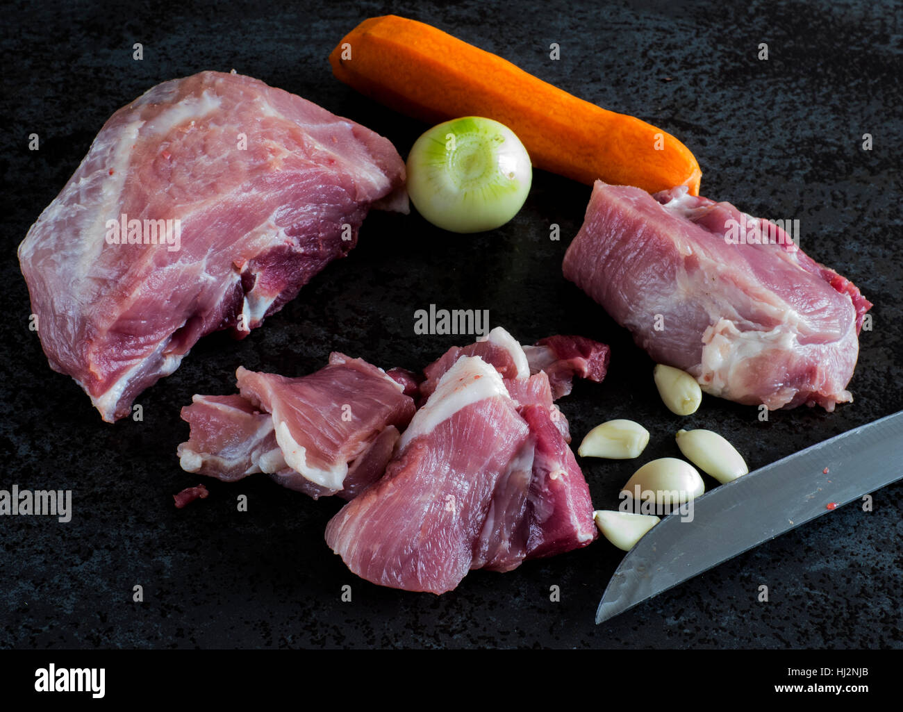 Raw joint pork meat, onion, garlic and carrot on dark stone background ...