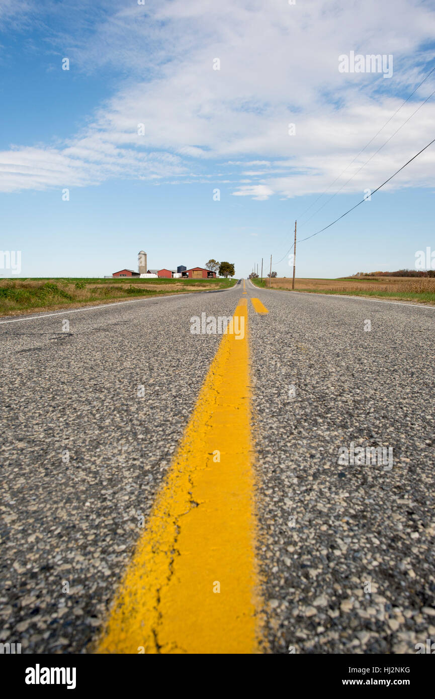 A paved country road with a red farmhouse and silo in the background ...