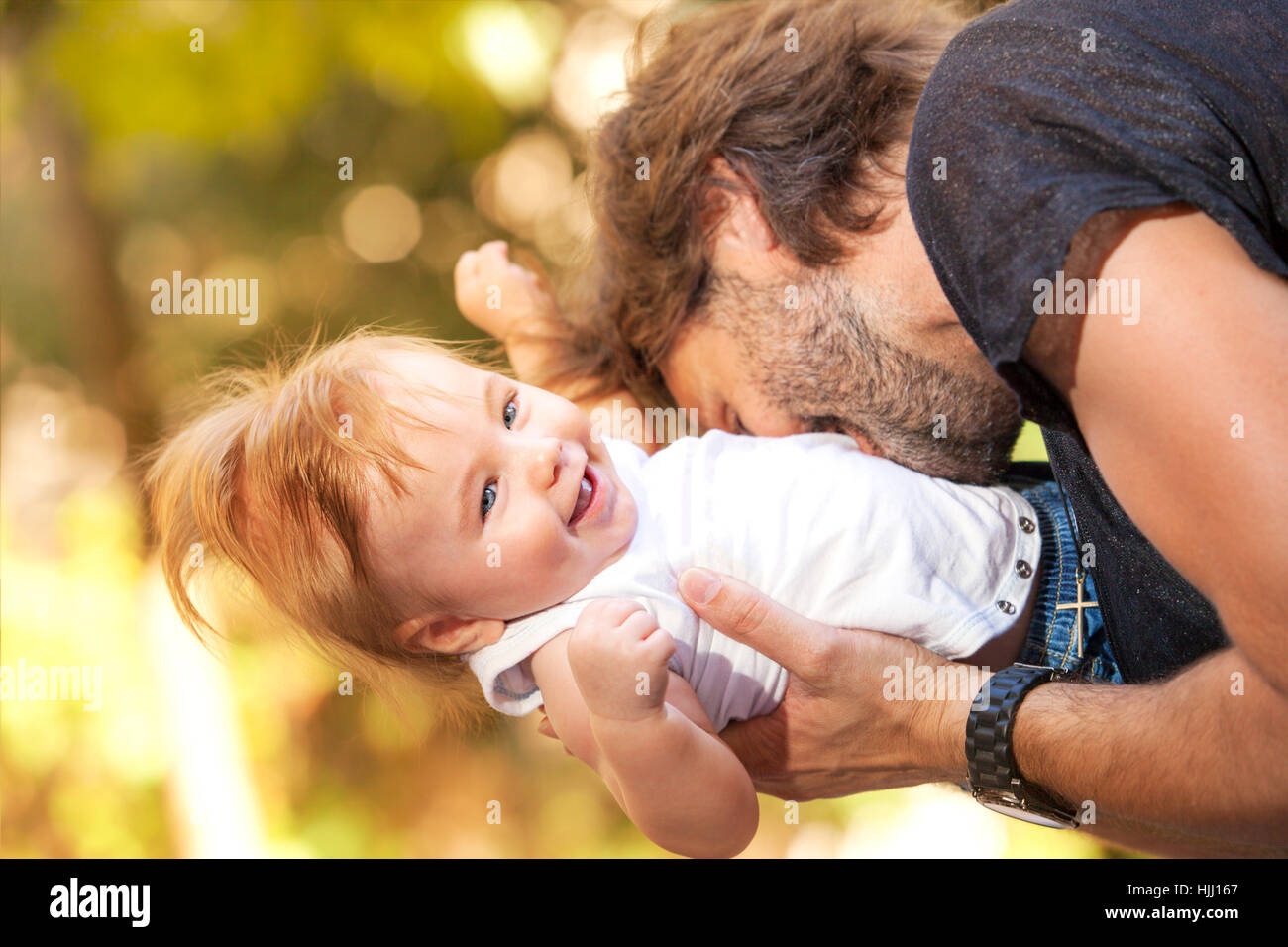 A cute little baby is laughing while his father is biting him playfully ...